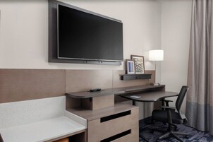 Quarto de hotel com televisão de tela plana na parede, uma escrivaninha com cadeira e uma pequena estante com luminária e molduras de quadros.