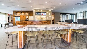 Uma área de bar moderna com um balcão comprido, várias telas e um espaço de estar com cadeiras de metal.