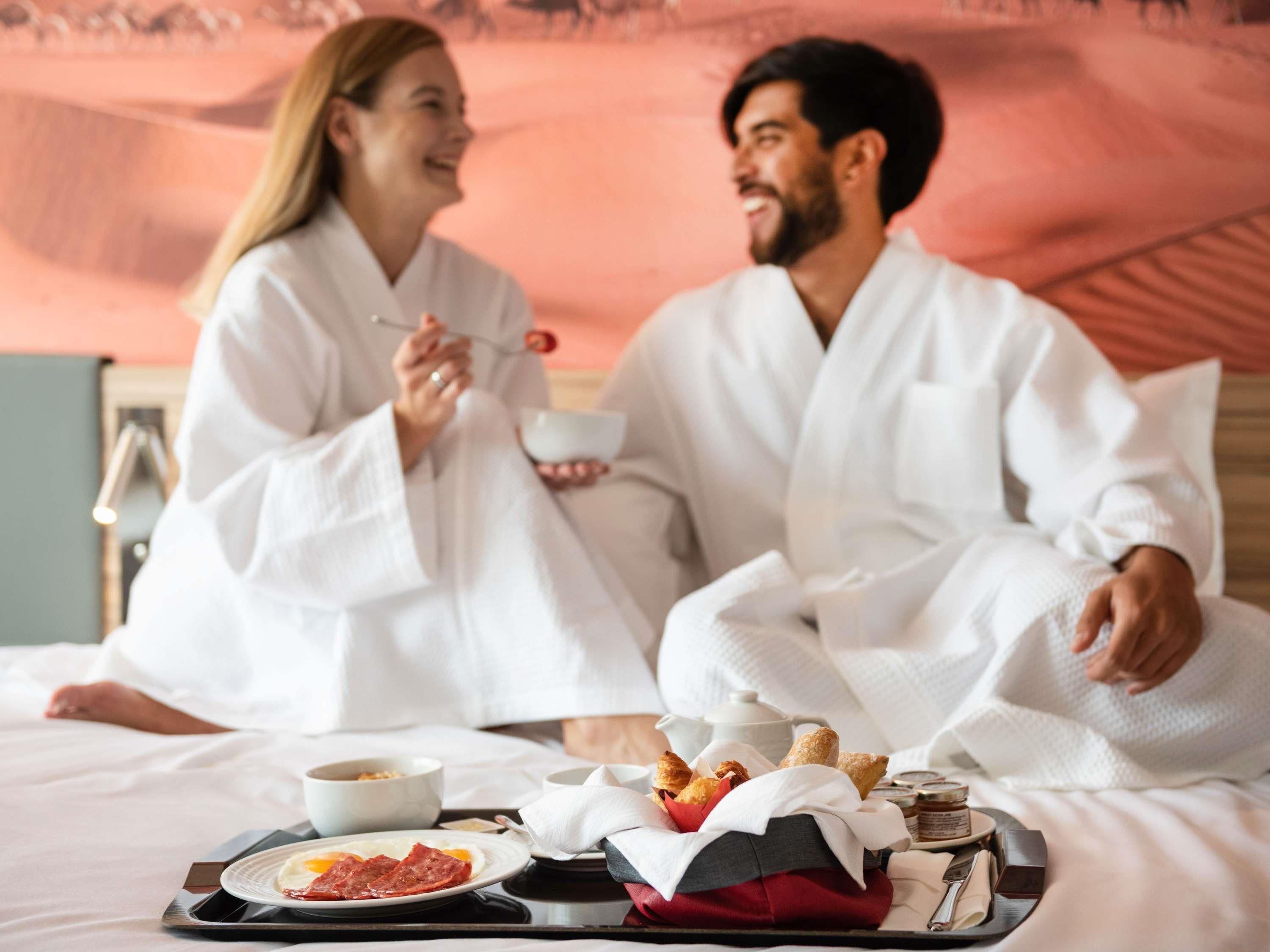 Um casal de vestes brancas sentado em uma cama com itens de café da manhã, incluindo ovos, doces e uma bandeja.