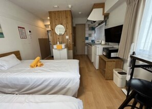 Quarto de hotel com duas camas, um pequeno espaço de cozinha, uma televisão e uma janela com cortinas.