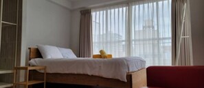 Uma cama de solteiro com estrutura de madeira e roupa de cama branca, uma cadeira vermelha e uma janela com cortinas leves.