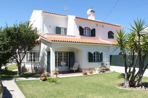 Uma casa de dois andares com telhado de telhas, venezianas verdes e varanda coberta com uma cadeira.