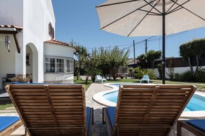 Uma área à beira da piscina com espreguiçadeiras de madeira e um grande guarda-sol.
