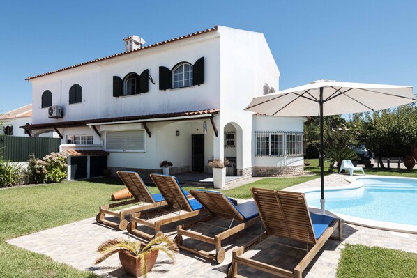 Um edifício branco com piscina, espreguiçadeiras de madeira e um guarda-sol.
