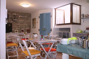 Uma área de refeições com cadeiras de madeira rústica, uma mesa com toalha verde e um espaço de cozinha com prateleiras e eletrodomésticos.
