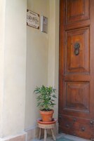 Uma porta de madeira com maçaneta de metal, uma planta em vaso sobre um banquinho, e uma placa com um número de telefone.