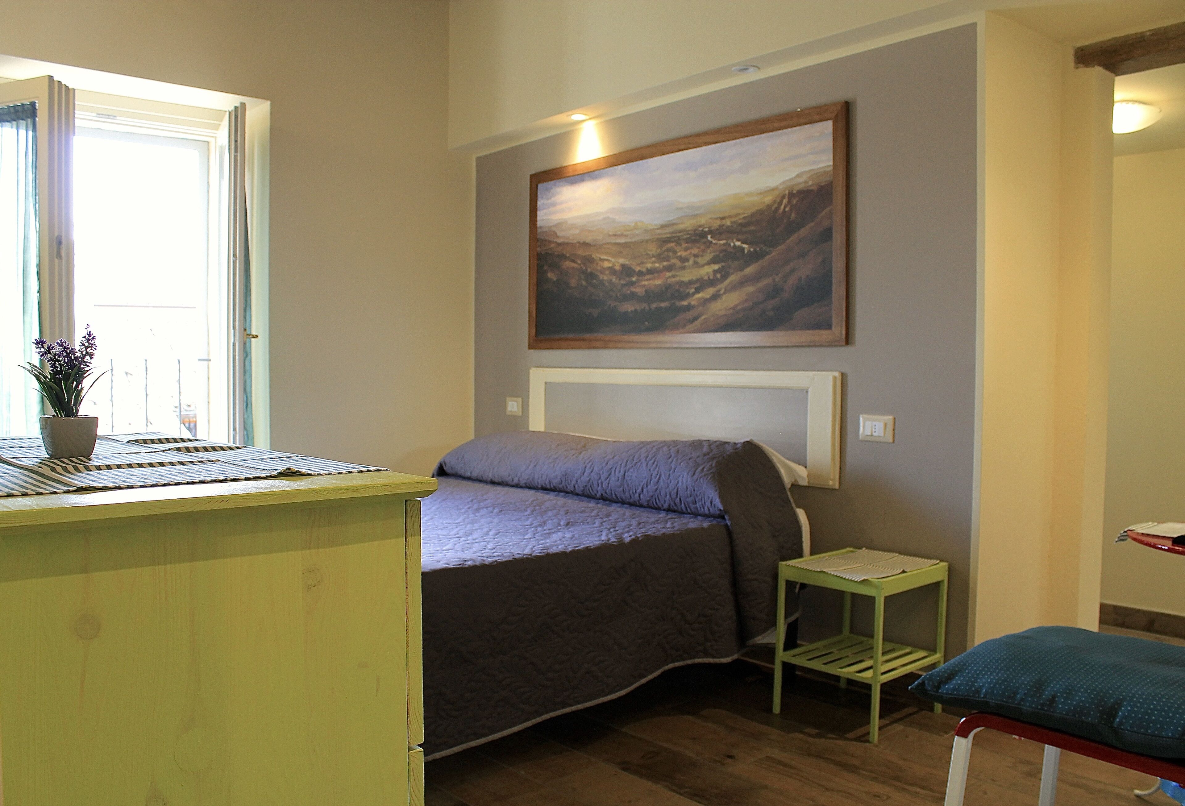 Quarto com cama, mesa de cabeceira pequena, janela com cortinas e um quadro na parede.