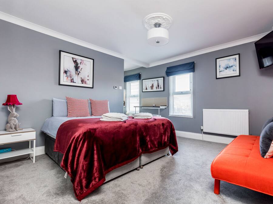 Quarto com cama, sofá vermelho, mesa de cabeceira branca e luminária vermelha.