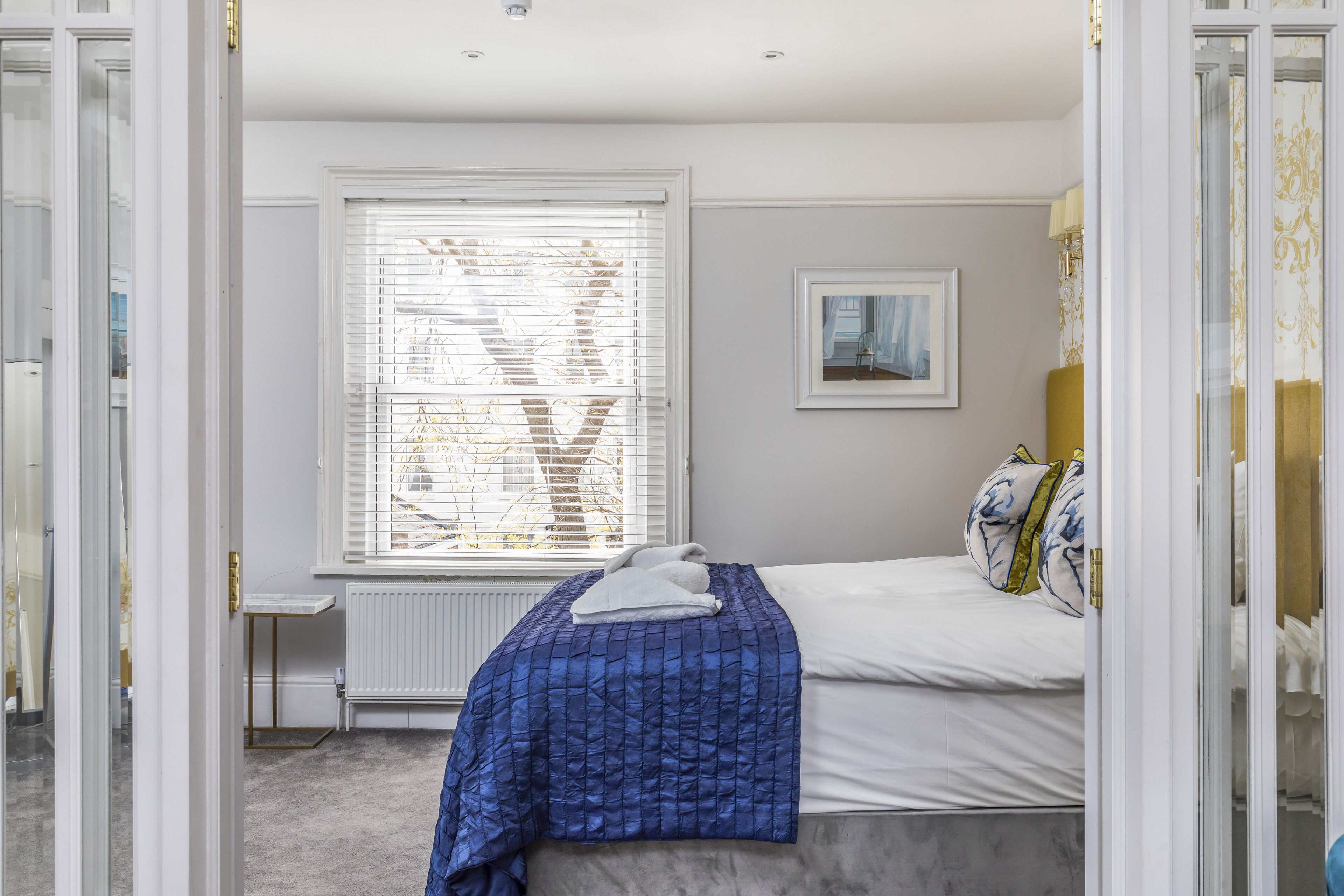 Quarto com cama, radiador, janela com persianas, mesa de cabeceira e um quadro na parede.