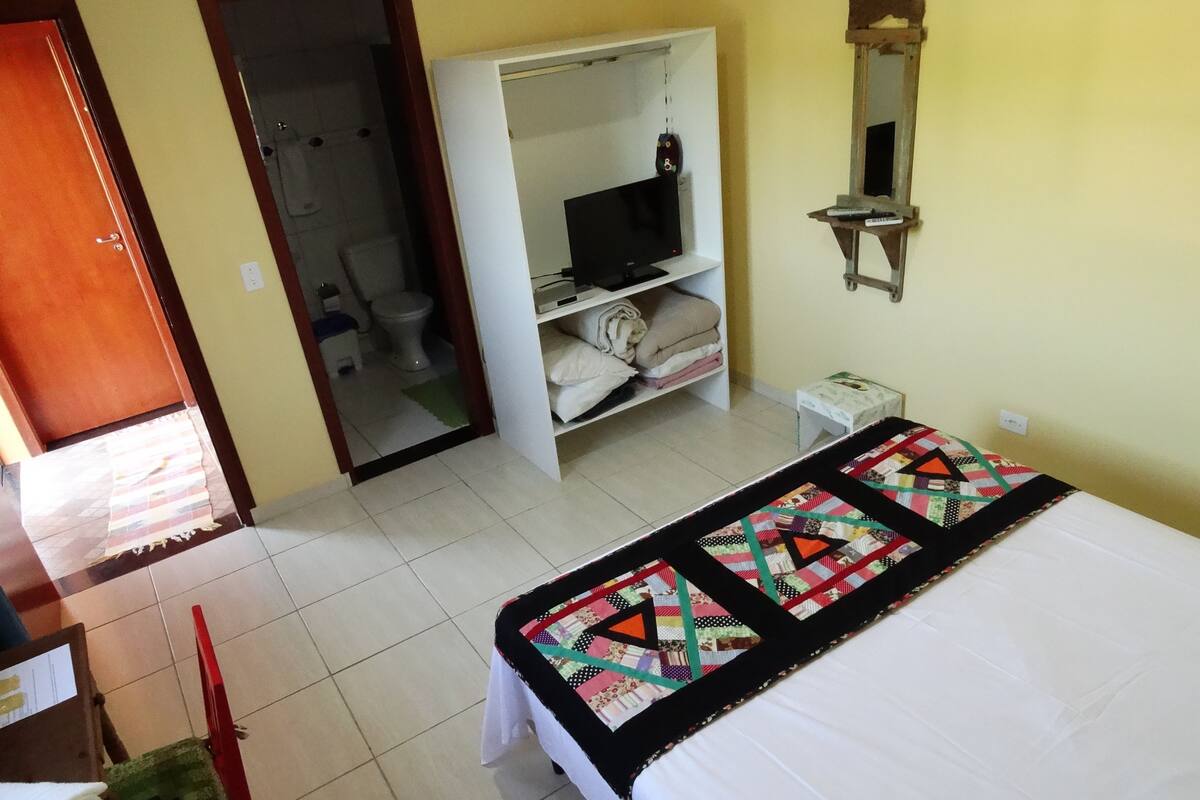 Quarto de hotel com cama, guarda-roupa, televisão, mesinha e espelho.