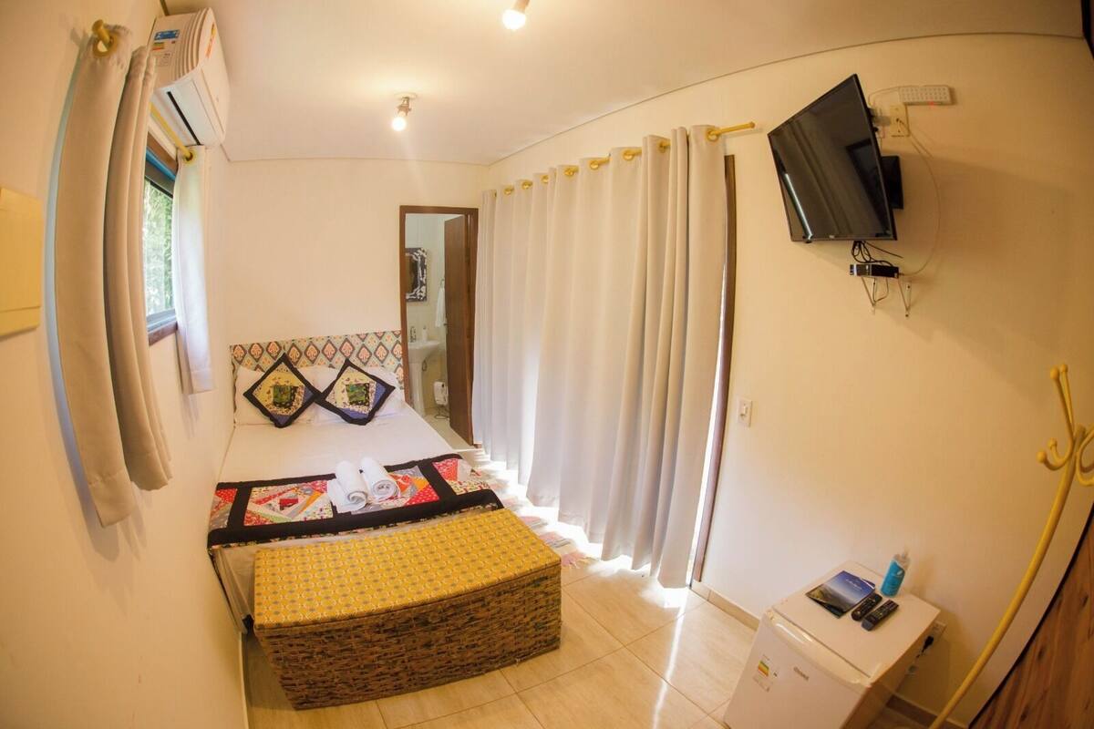 Quarto com cama, televisão, secretária pequena e janela com cortinas.