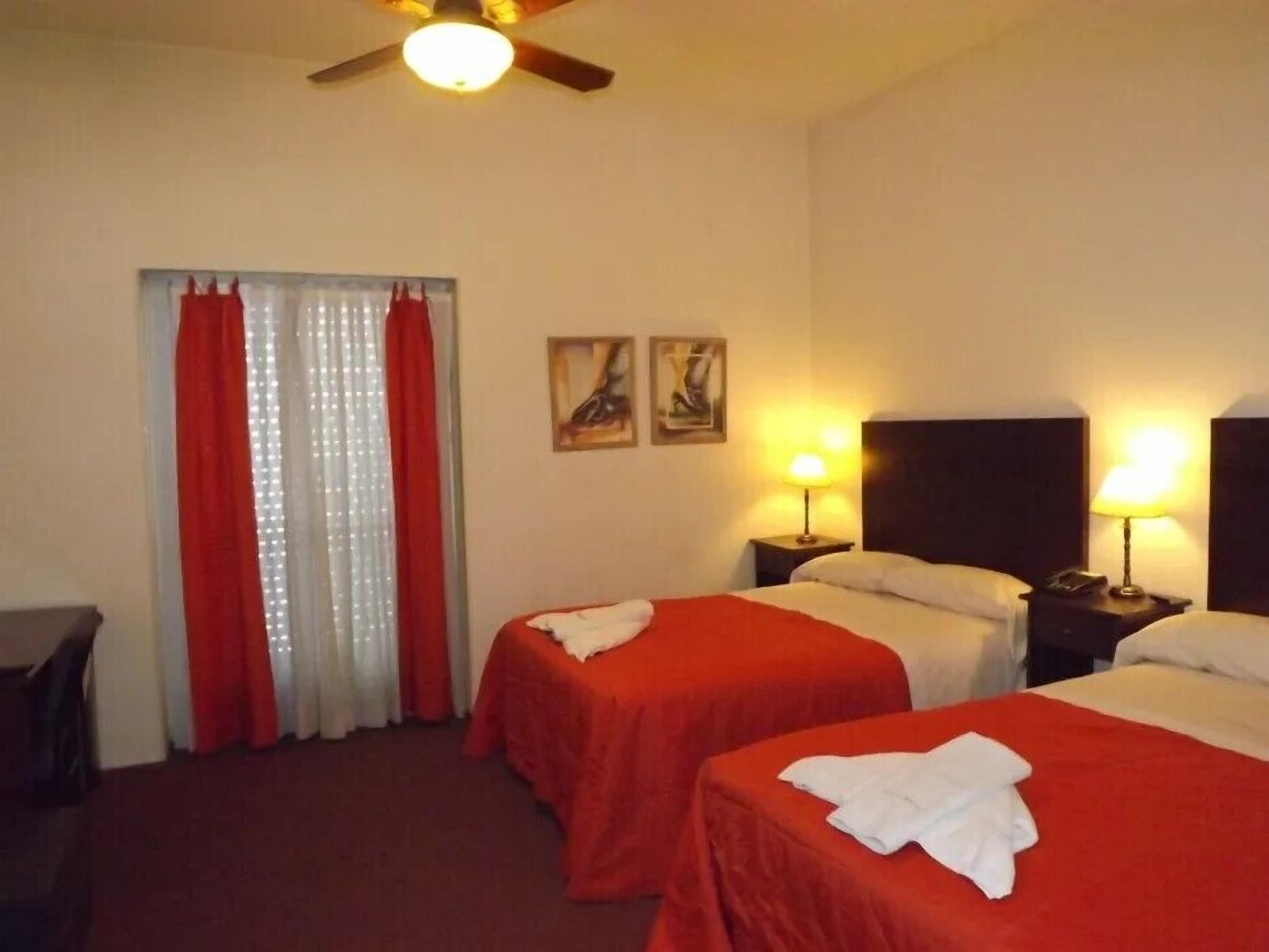 Quarto de hotel com duas camas, roupa de cama vermelha, um ventilador de teto e duas luminárias fixadas na parede.