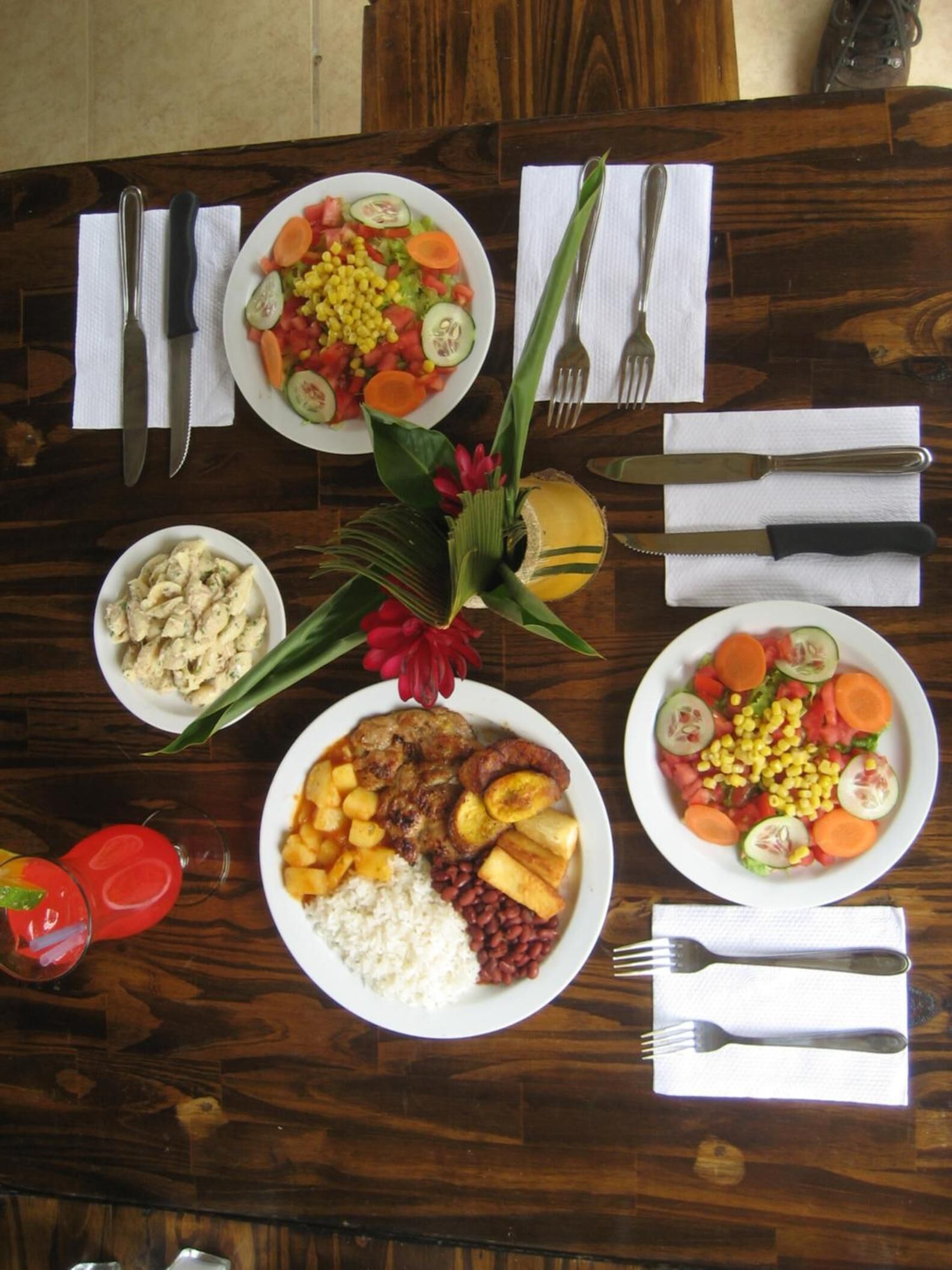 Uma mesa de jantar de madeira com vários pratos de comida, talheres e um arranjo floral decorativo.