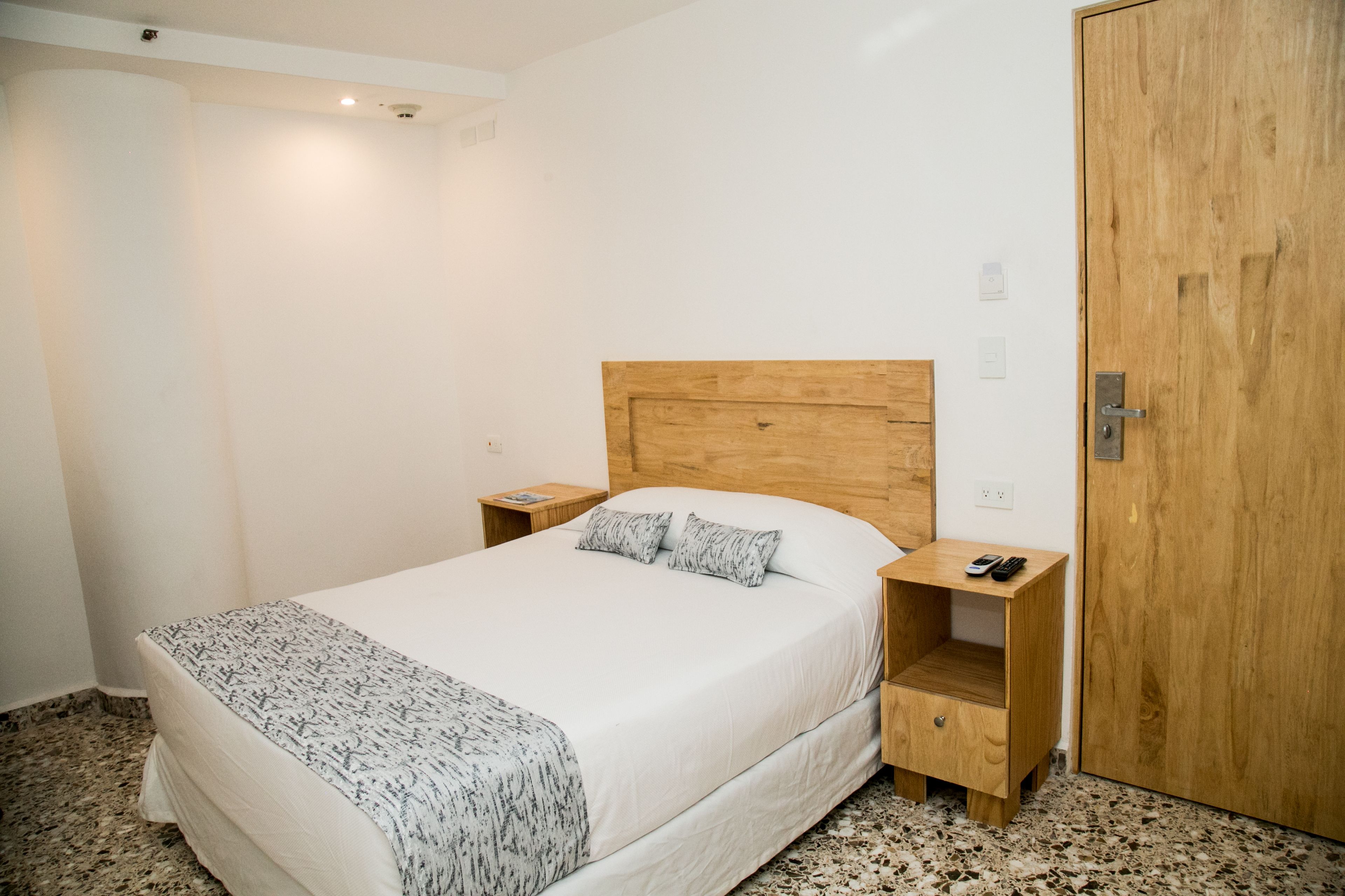 Quarto com cabeceira de madeira, cama com roupa de cama branca, mesa de cabeceira com controle remoto e guarda-roupa de madeira.