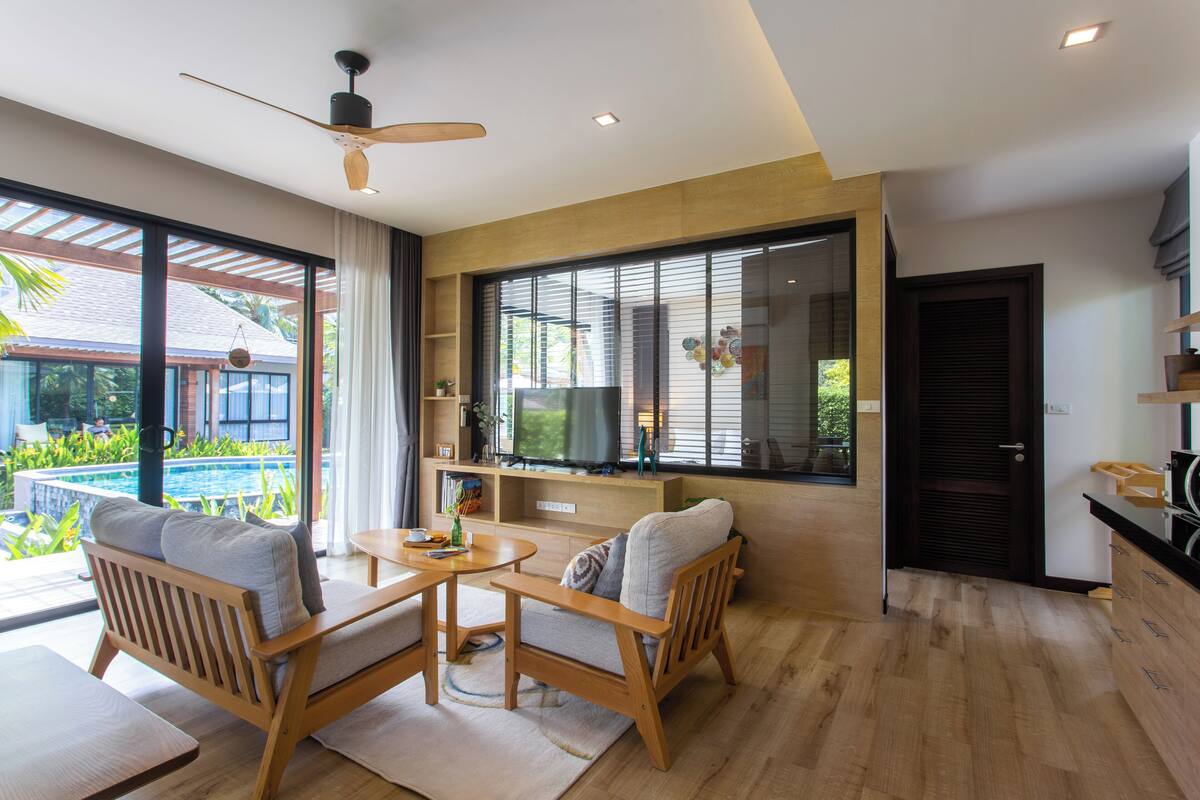 Uma sala de estar moderna com móveis de madeira, um ventilador de teto e uma grande janela com vista para uma piscina.