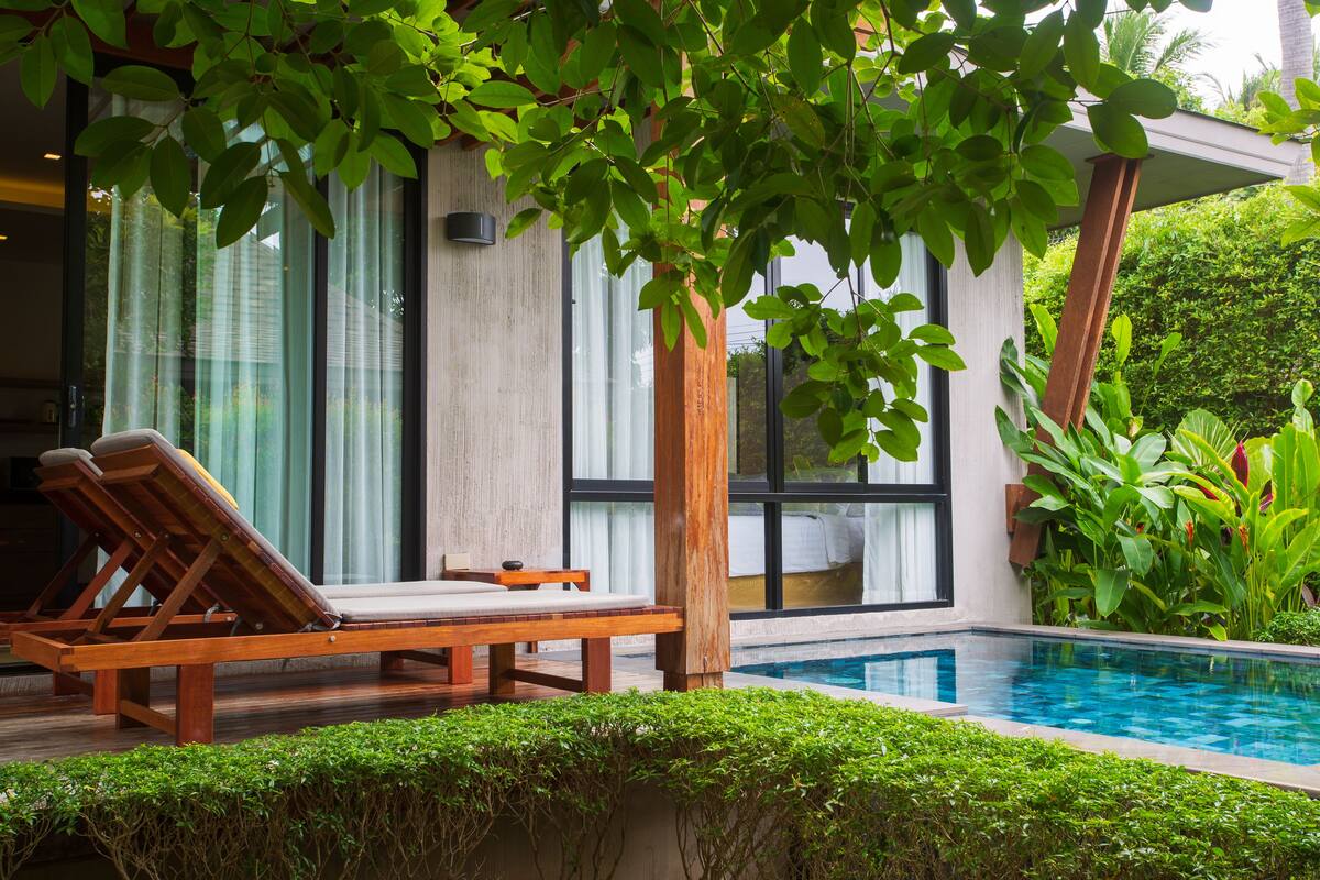 Uma área na piscina com espreguiçadeira de madeira e uma mesinha, cercada por vegetação exuberante e um edifício moderno com janelas amplas.