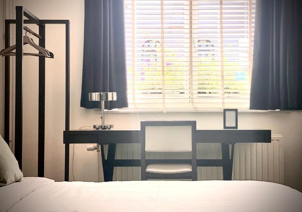 Quarto de hotel com cama, escrivaninha com cadeira, janela com cortinas e radiador.