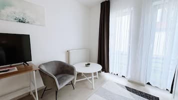 Uma sala de estar moderna e minimalista, com televisão de tela plana, uma poltrona cinza, uma mesa pequena branca e uma grande janela com cortinas leves.