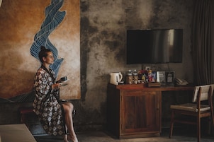 Uma pessoa sentada num sofá em um quarto com uma televisão de tela plana grande fixada na parede, um armário de madeira com diversos objetos e um design de parede estampado.