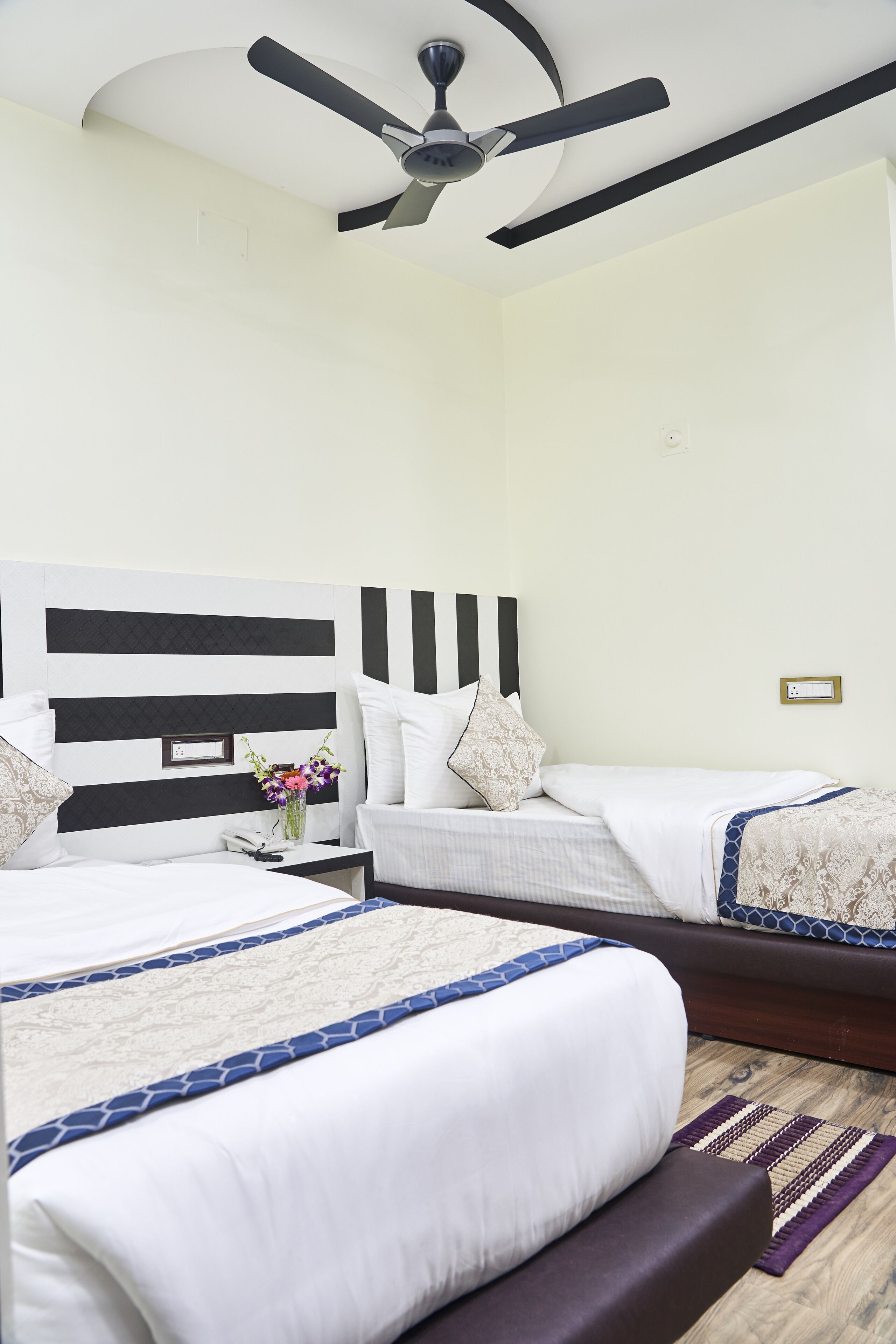 Quarto de hotel com duas camas, um ventilador de teto, uma mesa de cabeceira com um vaso de flores e piso de madeira.