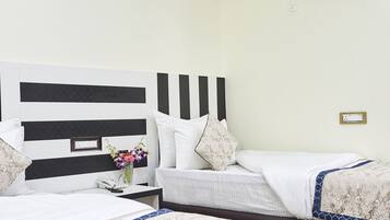Quarto de hotel com duas camas, um ventilador de teto, uma mesa de cabeceira com um vaso de flores e piso de madeira.
