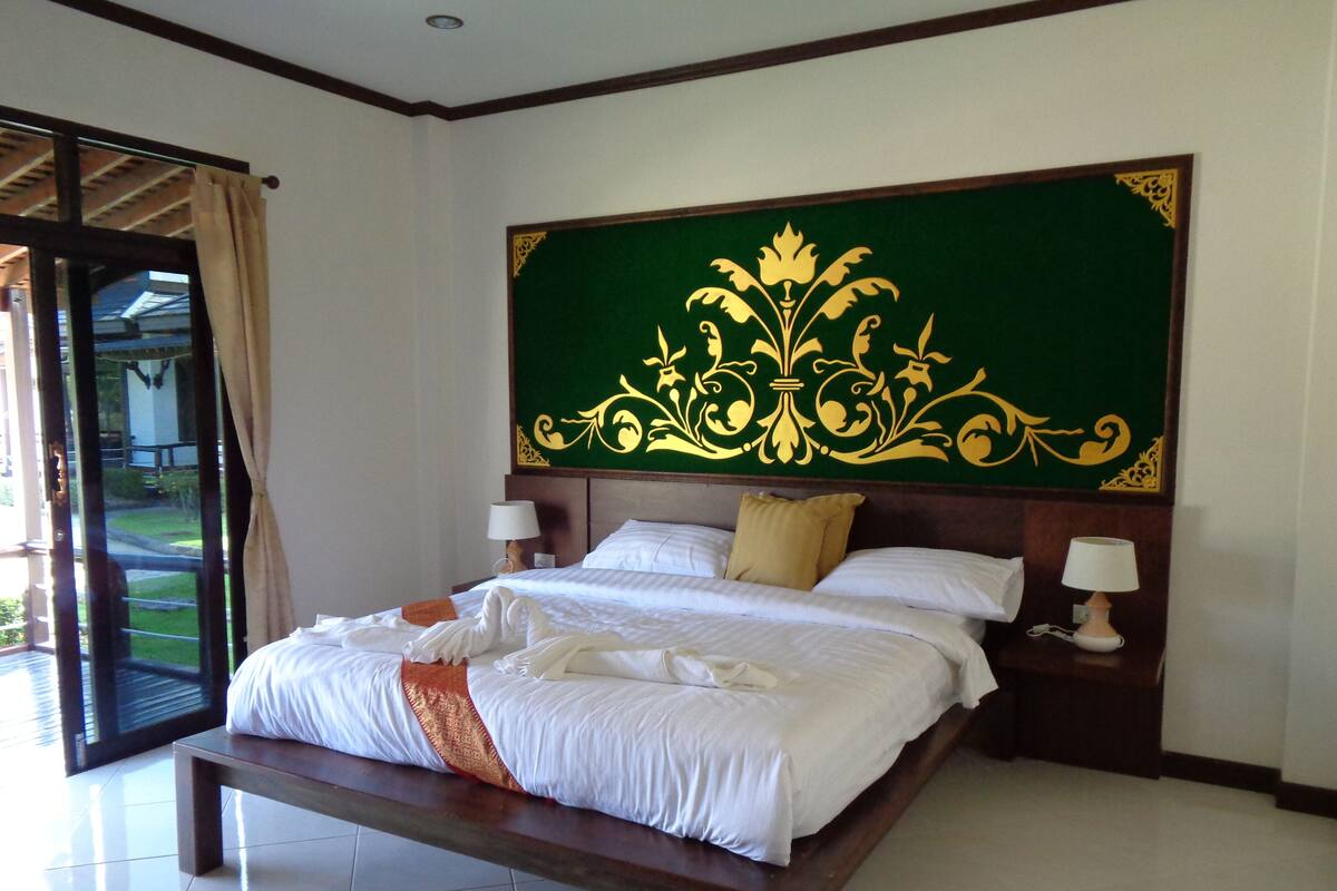 Uma cama grande com lençóis brancos, duas mesas de cabeceira com luminárias, um painel de cabeceira decorativo verde e vista para um jardim por uma porta de vidro.