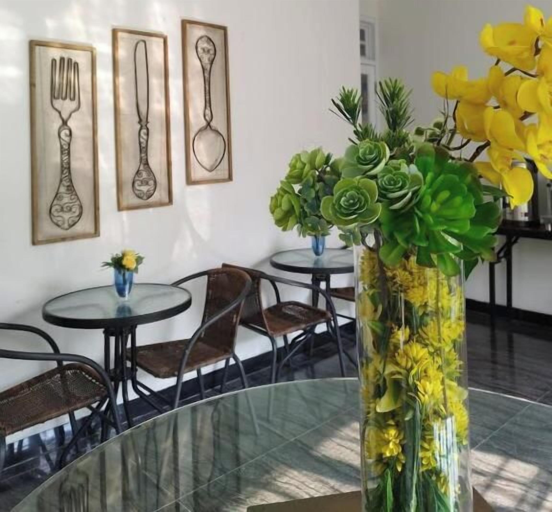 Uma área de refeições com mesas redondas, cadeiras e uma mesa com tampo de vidro. Há quadros nas paredes acima das mesas.