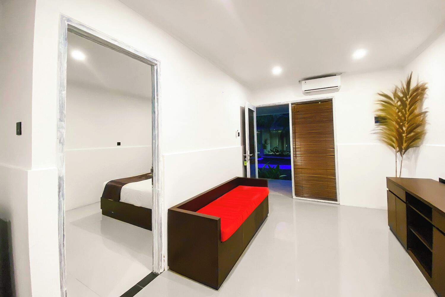 Quarto de hotel moderno com sofá de destaque vermelho, um espelho grande e vista para área de piscina externa.
