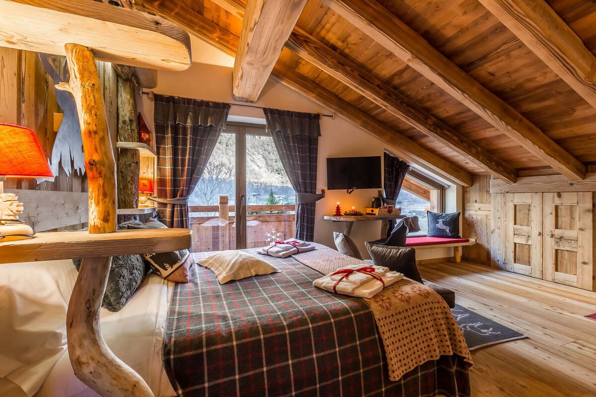 Um quarto aconchegante com teto de madeira, cama com roupa de cama xadrez, mesa de madeira com abajur e janela com cortinas.