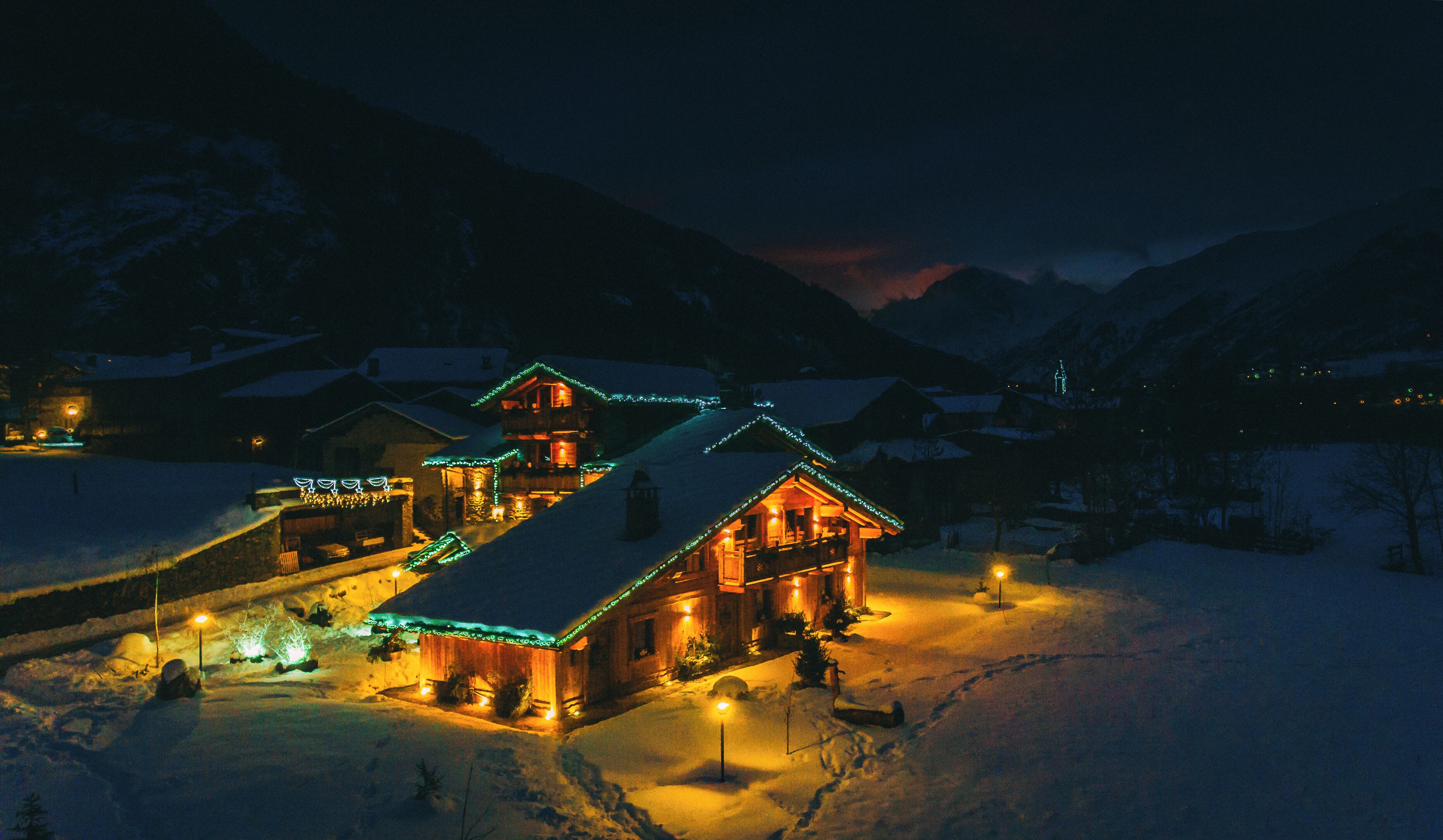 Uma pousada coberta de neve com vários edifícios, iluminada por luzes quentes, rodeada por montanhas.