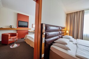 Quarto de hotel com cama, televisão na parede, poltrona vermelha e uma mesinha de cabeceira com abajur.