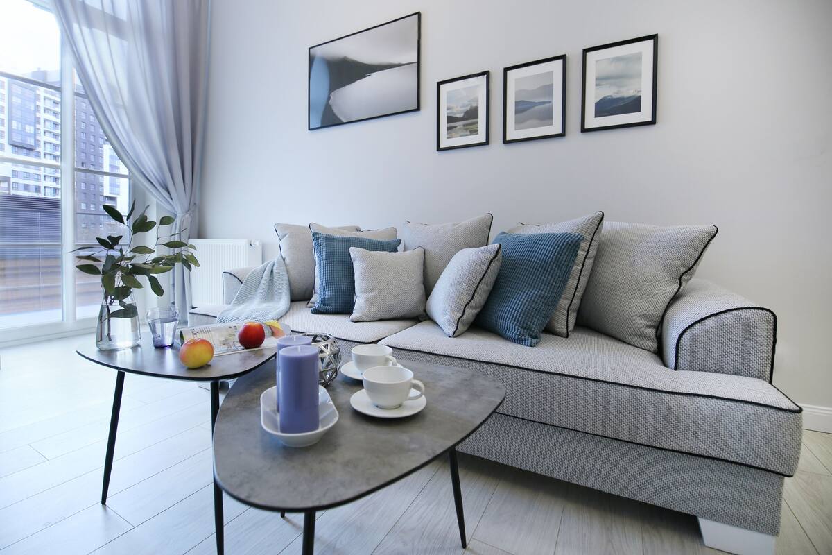 Uma sala de estar moderna com um sofá cinzento, almofadas azuis e brancas, uma mesa de centro com um vaso e frutas, e quadros nas paredes.