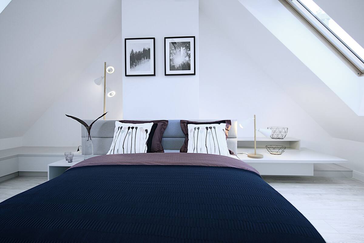 Um quarto moderno com teto inclinado, uma cama com colcha azul escura, duas imagens emolduradas na parede e um claraboia.