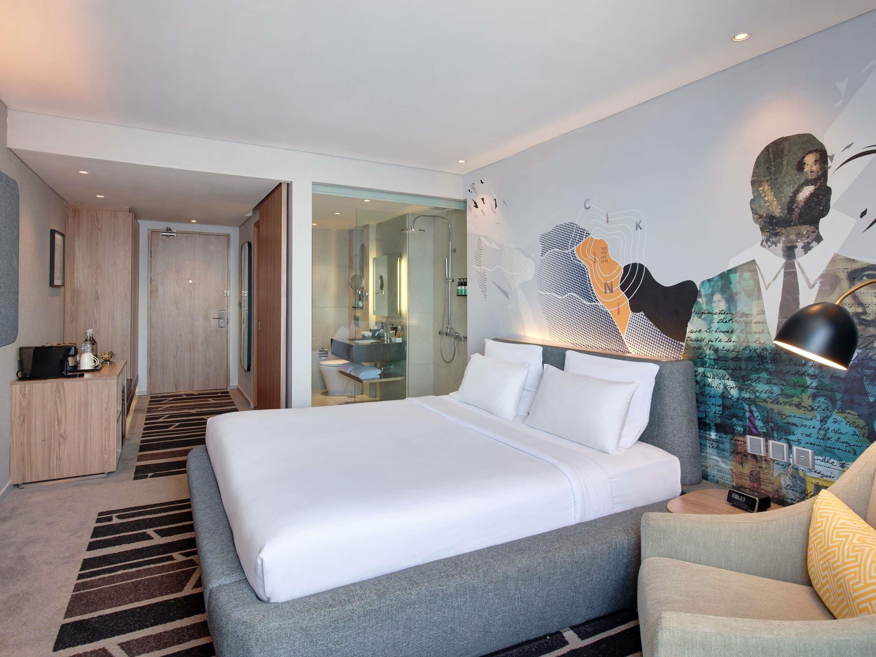 Quarto de hotel moderno com uma cama grande, uma escrivaninha e um banheiro visível através de um painel de vidro.