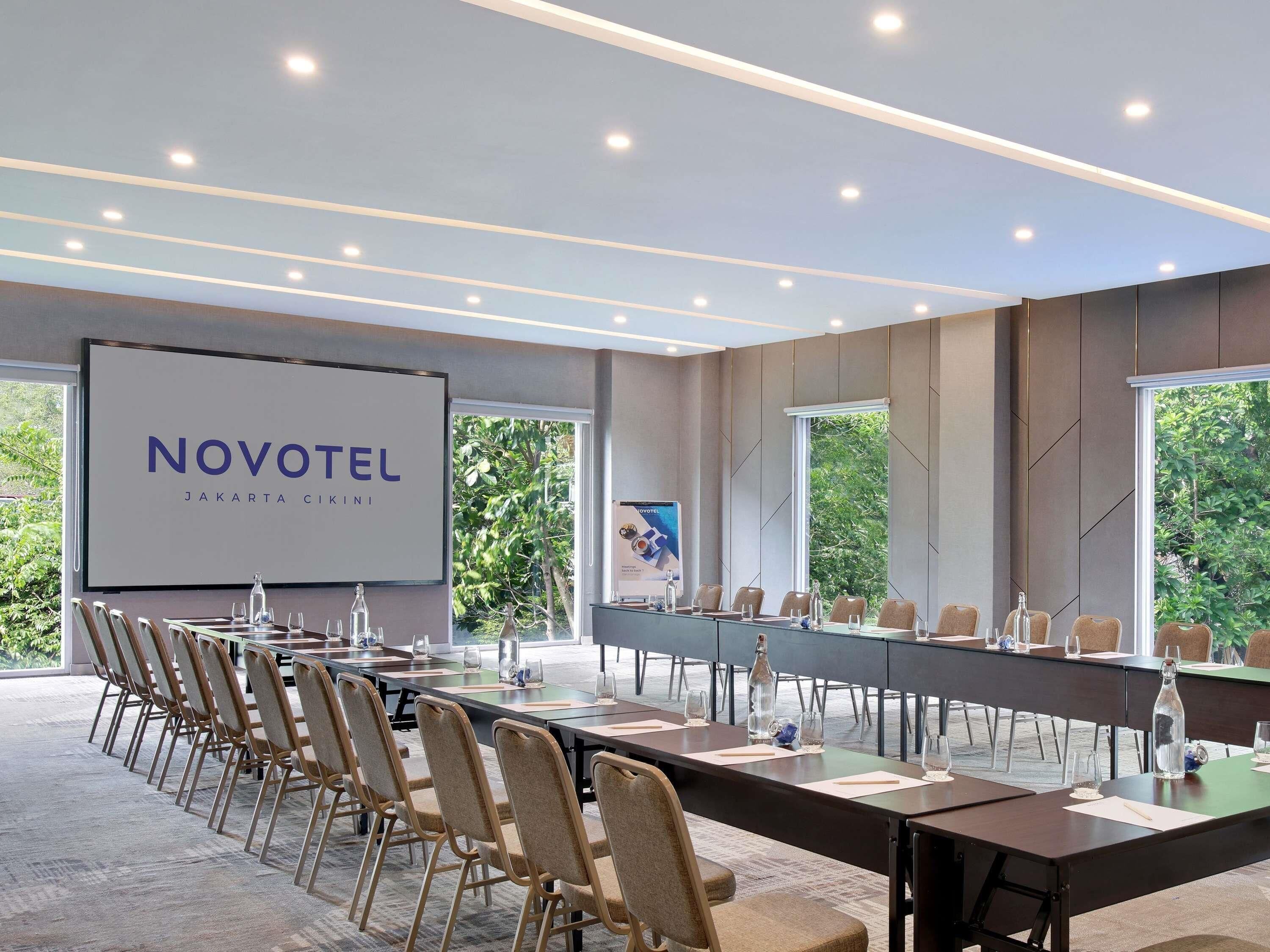 Uma sala de conferências com uma mesa comprida, cadeiras, uma tela de projetor exibindo 'NOVOTEL JAKARTA CIKINI' e grandes janelas com vista para o verde.