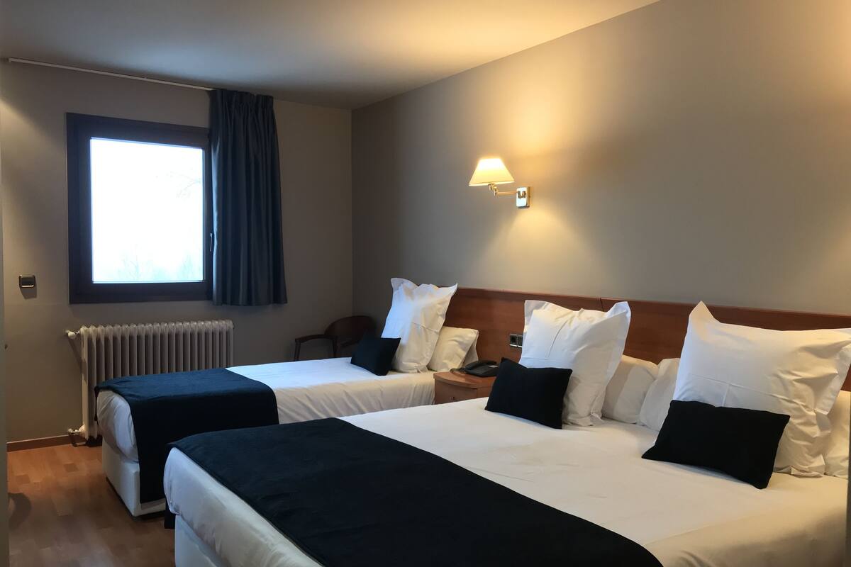 Quarto de hotel com duas camas, janela com cortinas, mesa de cabeceira com abajur e radiador.