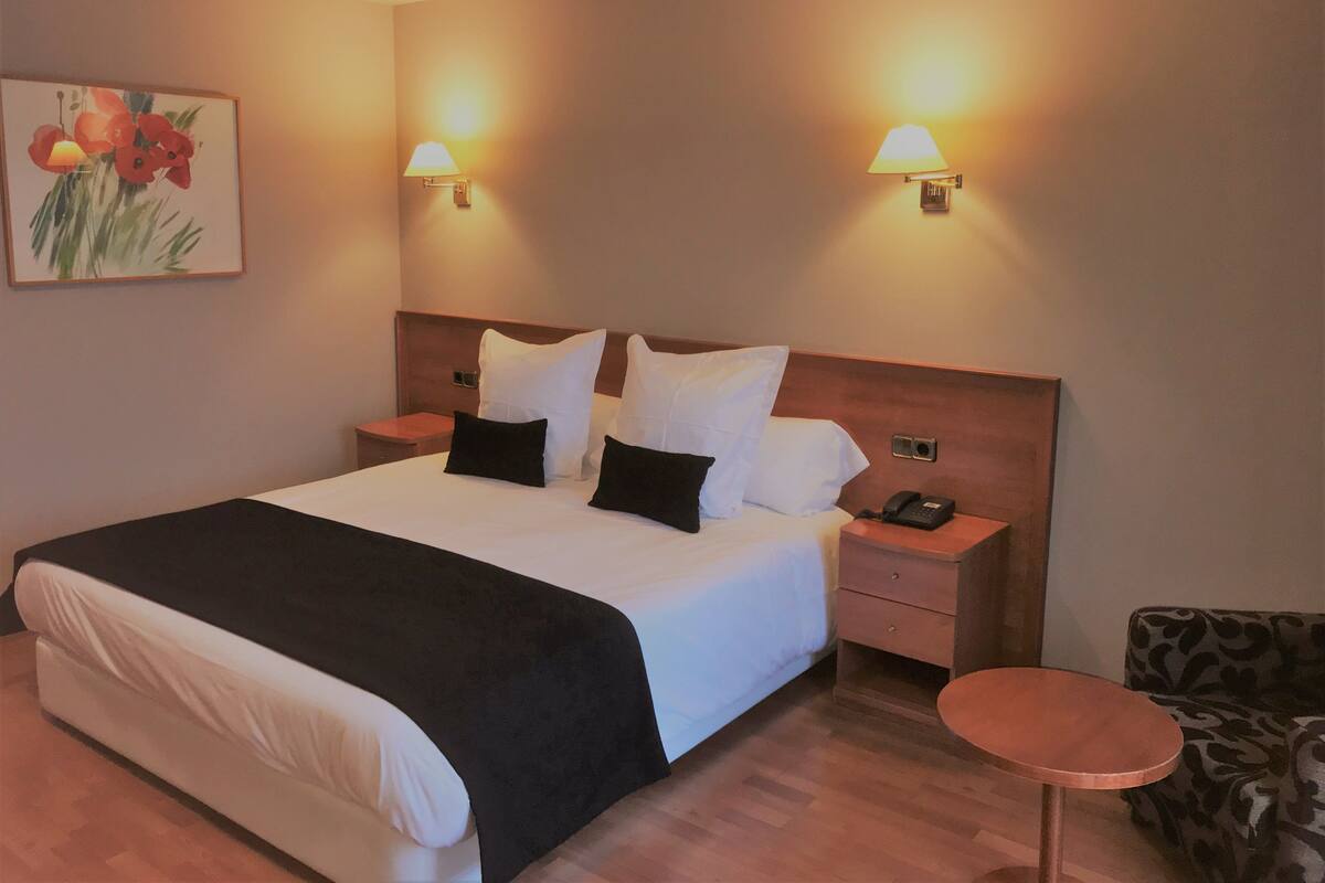 Quarto de hotel com uma cama grande, duas mesas de cabeceira, uma mesinha redonda, uma poltrona estampada e duas luminárias fixadas na parede.