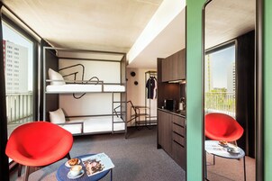 Quarto de hotel compacto com beliches, cozinha compacta, área de jantar com uma cadeira vermelha e vista para a cidade.