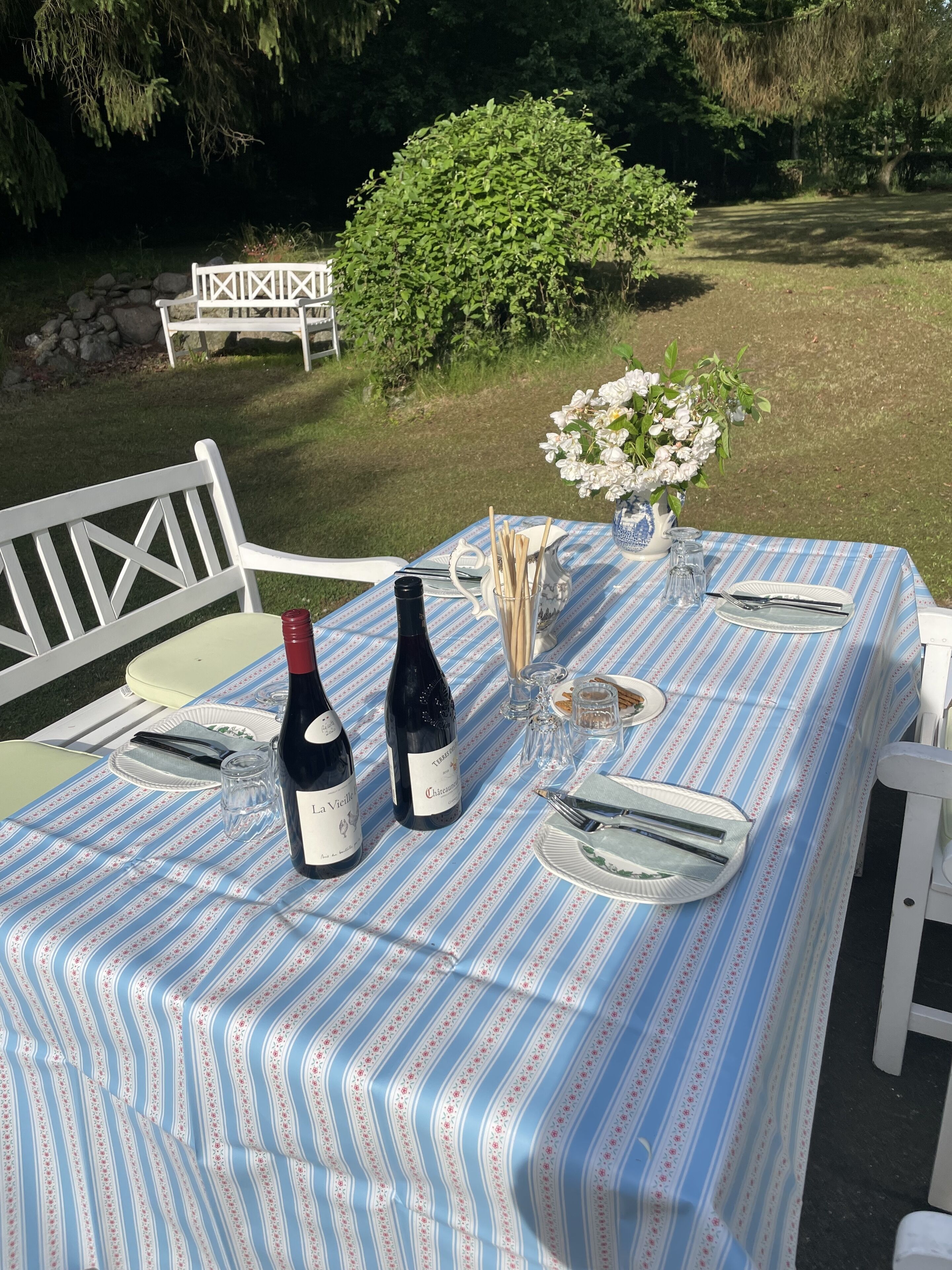 Mesa de jantar ao ar livre posta com toalha listrada, duas garrafas de vinho, copos e um vaso de flores.