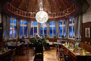 Uma sala de jantar com mesas e cadeiras de madeira, janelas amplas e um candelabro.