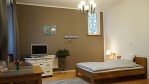 Quarto com cama de madeira, escrivaninha com computador, televisão, planta e janela com vista.