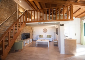 Ampla sala de estar com escada de madeira, sofá branco, TV de tela plana e parede de destaque em tijolo.