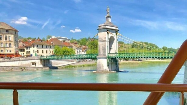 Uma mesa com um copo de água e um guardanapo, uma cadeira de vime e vista de um rio com uma ponte e edifícios ao fundo.