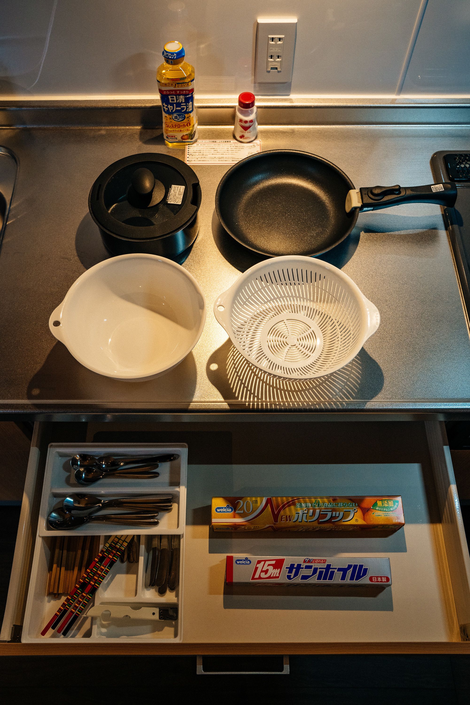 Uma bancada de cozinha com uma frigideira, uma tigela branca, um escorredor e uma garrafa de óleo de cozinha. Abaixo da bancada, há uma gaveta com utensílios e uma caixa de Velcro.