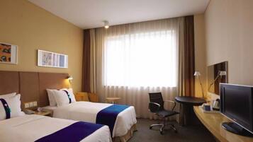 Quarto de hotel com duas camas, uma mesa de trabalho com televisão, uma cadeira e uma janela com cortinas.