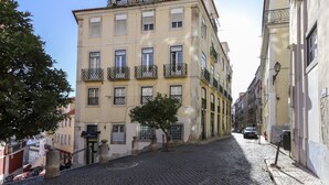 Uma rua de paralelepípedos estreita, ladeada por edifícios com várias andares e varandas, com uma árvore no centro.