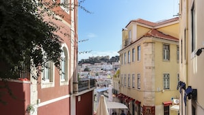 Uma rua estreita com edifícios de ambos os lados, um prédio vermelho à esquerda e um prédio amarelo à direita. Há mesas e cadeiras ao ar livre sob guarda-sóis, e algumas pessoas caminhando.