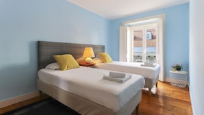 Quarto de hotel com duas camas, uma janela com vista para um edifício e uma mesinha de cabeceira com uma luminária.