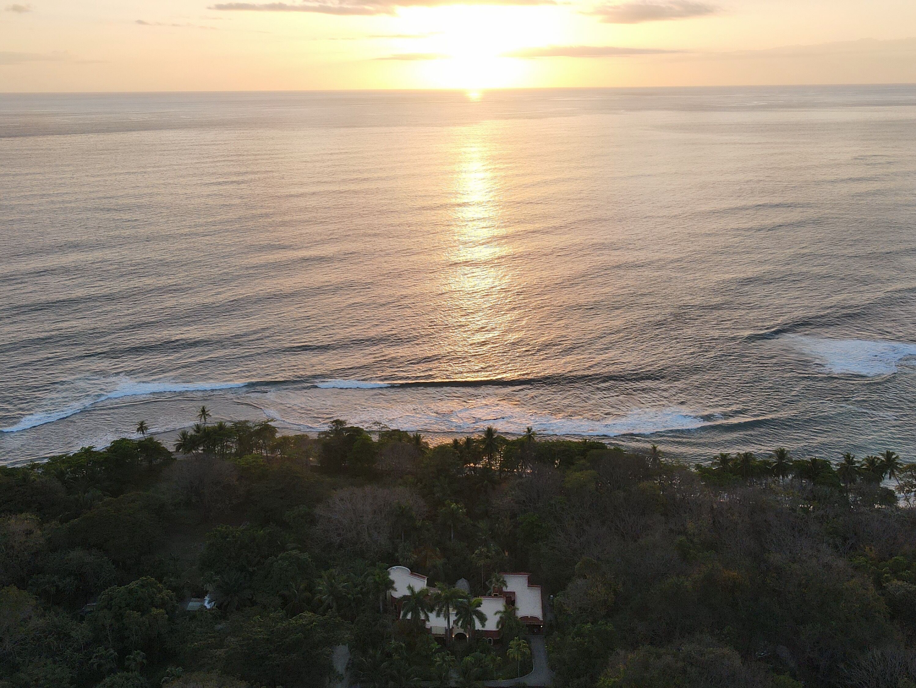 Vista costeira com pôr do sol sobre o oceano, uma praia com palmeiras e um pequeno edifício em primeiro plano.