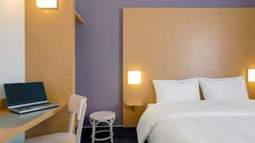 Quarto de hotel com uma cama, uma escrivaninha com um laptop, uma cadeira e duas luzes fixadas na parede.